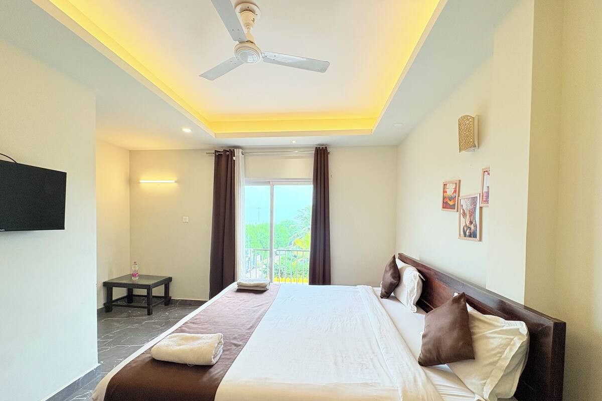 Quarto de hotel com cama, ventilador de teto, televisão, mesinha e vista para o verde pela janela.