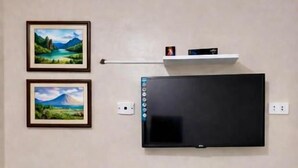 Quarto com televisão de tela plana na parede, uma secretária branca, duas cadeiras de madeira e um frigorífico pequeno.