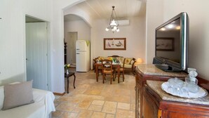 Uma sala de estar acolhedora, com um sofá, uma mesa de jantar com cadeiras, uma TV sobre um aparador de madeira e um lustre.
