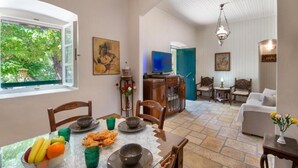 Uma área de refeições com uma mesa posta para dois, uma televisão ao fundo e vista para o exterior pela janela.