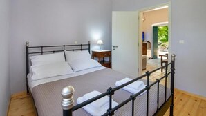 Uma cama de solteiro com estrutura de metal, uma mesa de cabeceira com abajur e uma porta que dá para uma área externa.