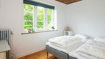 Quarto com chão de madeira, cama com roupa de cama branca, janela vista para vegetação e radiador.