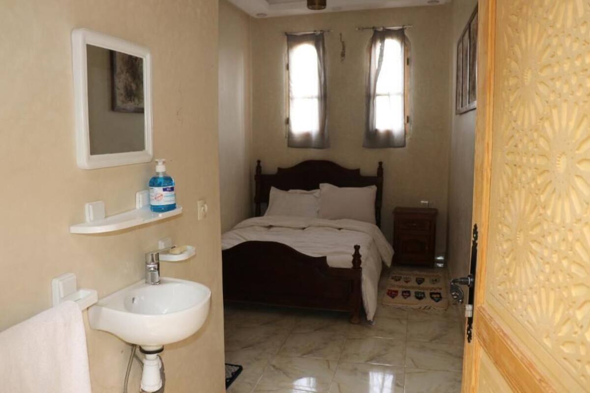 Quarto com cama, mesa de cabeceira, espelho, prateleira com artigos de higiene e janela com cortinas.