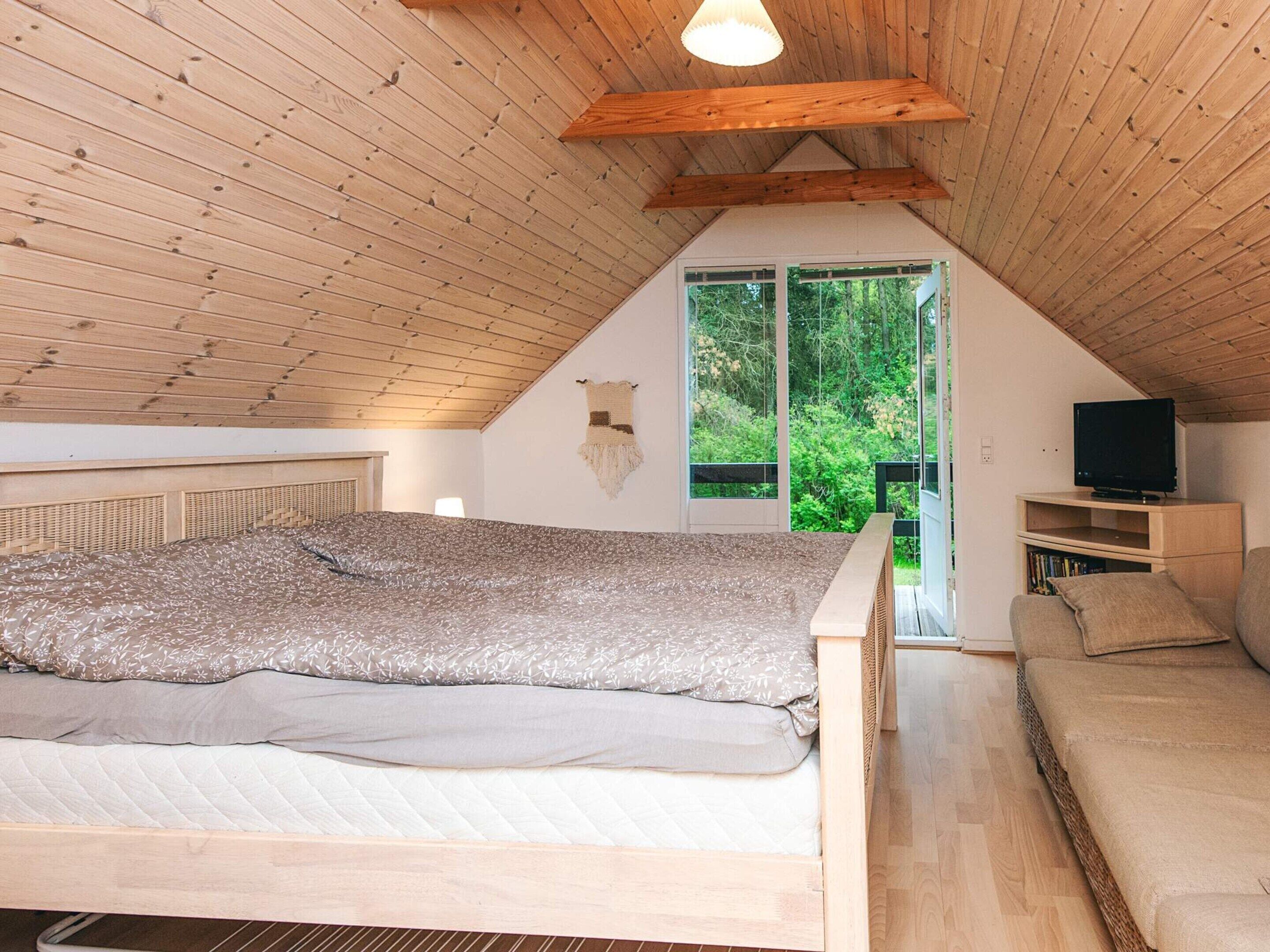 Quarto com teto inclinado, uma cama, um sofá, uma televisão e vista para o verde lá fora.