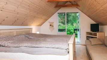 Quarto com teto inclinado, uma cama, um sofá, uma televisão e vista para o verde lá fora.