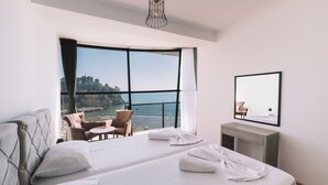 Quarto de hotel com uma cama grande, uma cadeira, uma mesa pequena, uma mesinha de cabeceira e vista para o mar.