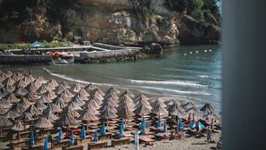Uma praia com fileiras de guarda-sóis de palha e espreguiçadeiras de madeira, um penhasco rochoso ao fundo e um mar calmo.