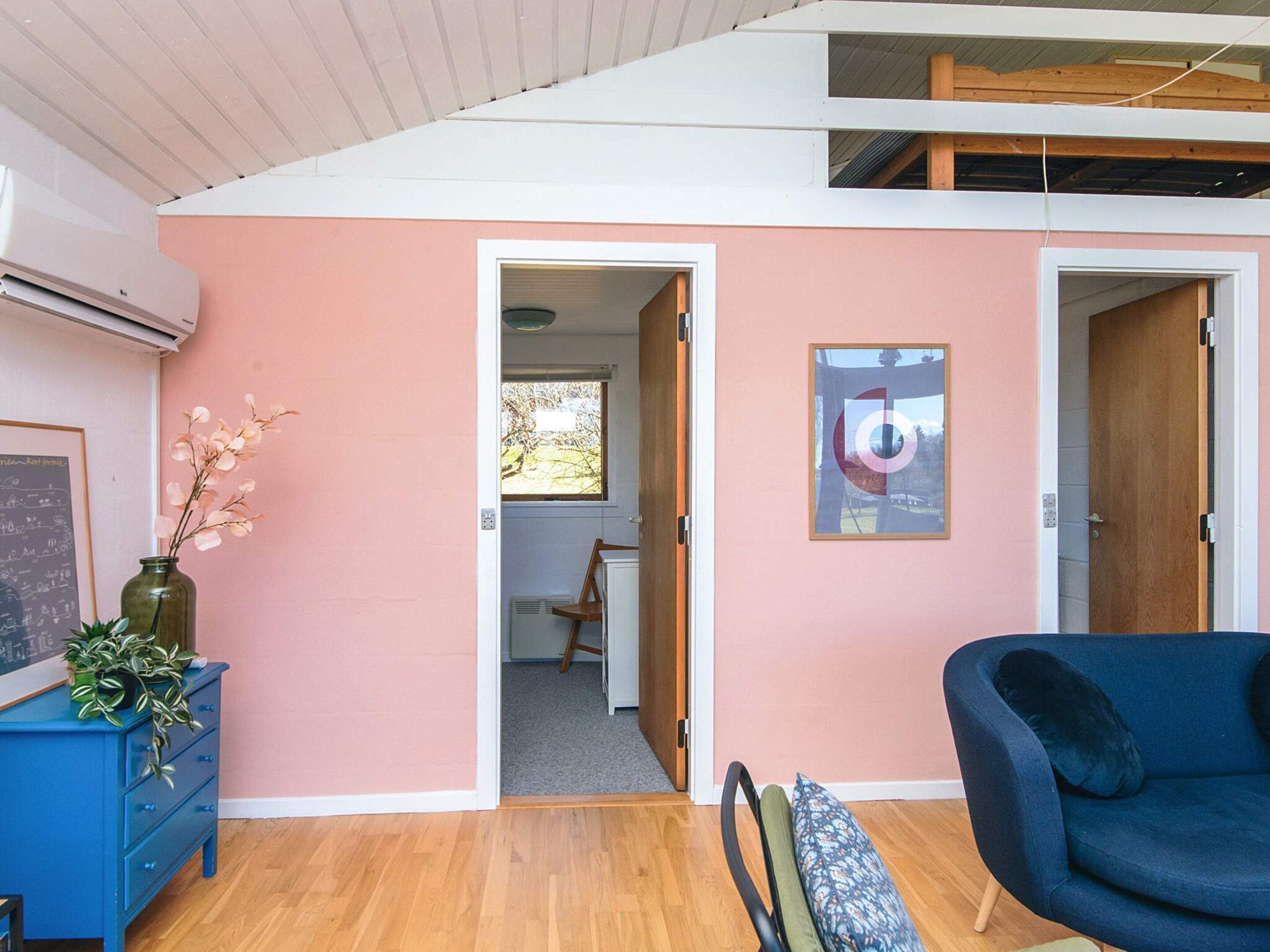 Quarto com parede rosa, cadeira azul, chão de madeira e porta que dá para outro cômodo.
