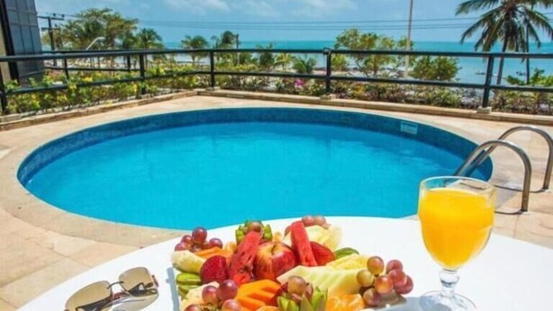 Cenário à beira da piscina com uma bandeja de frutas, um copo de suco de laranja e óculos de sol sobre uma mesa.