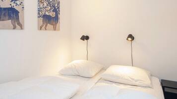 Uma cama com roupa de cama branca, duas luminárias pretas fixadas na parede e dois quadros na parede.