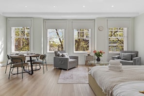 Quarto espaçoso com uma cama grande, dois sofás cinzentos, uma mesa de jantar com cadeiras e três janelas amplas com vista para árvores.