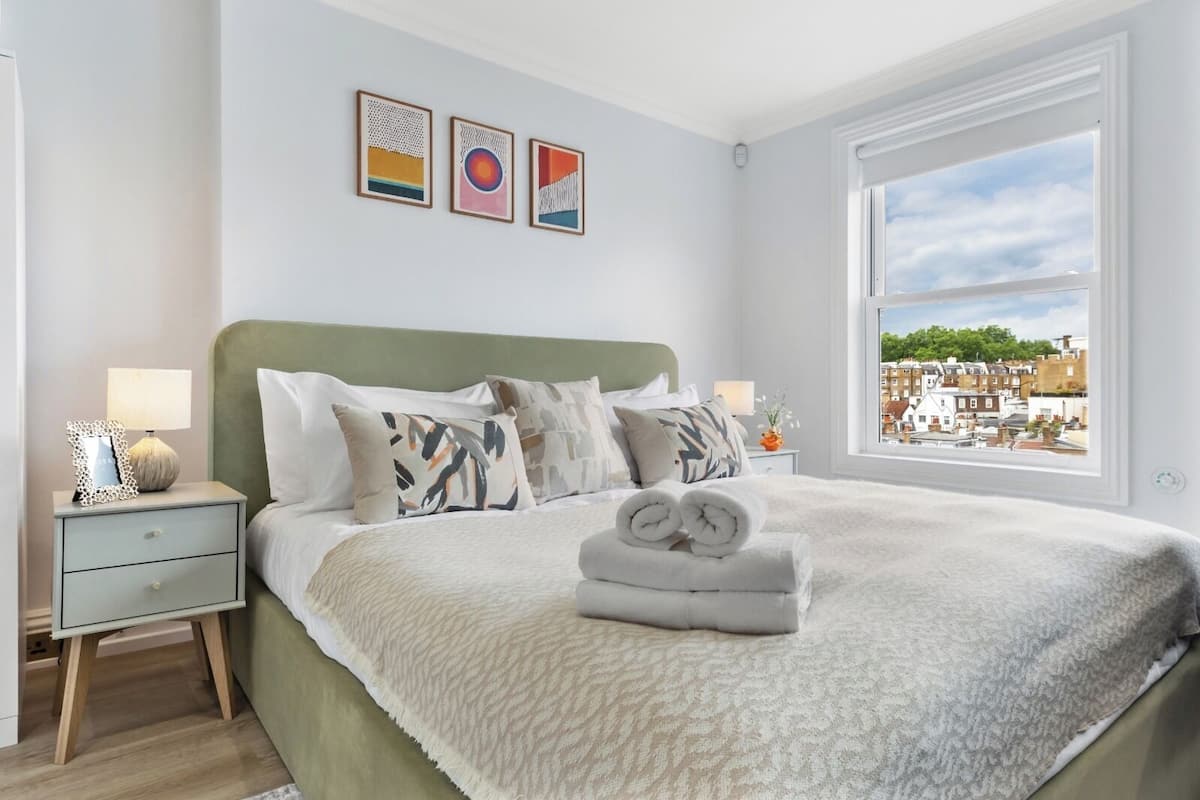 Quarto com cama, almofadas, mesa de cabeceira, luminária e janela com vista da cidade.