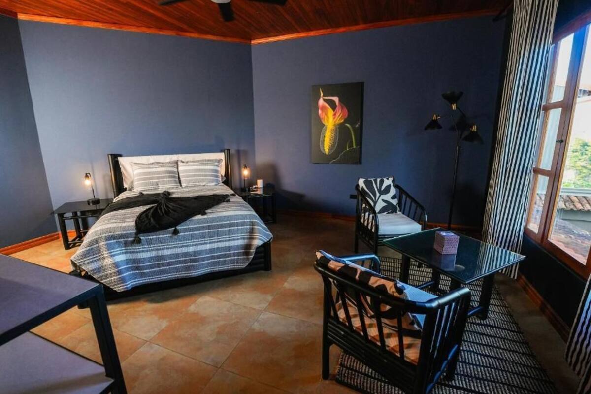 Quarto com cama, mesa de cabeceira, cadeira, pequena mesa e janela.