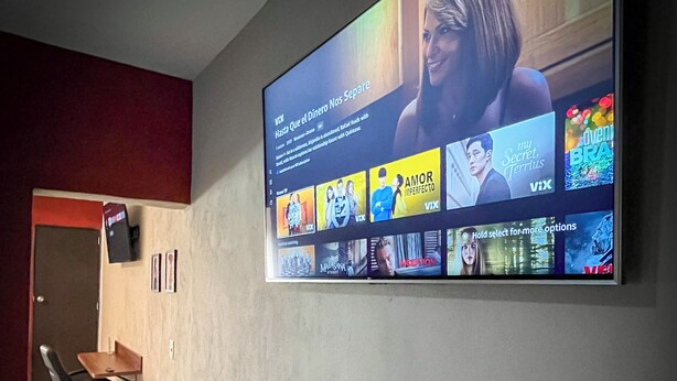 Uma sala de conferências com uma tela grande exibindo uma interface de videoconferência, um banco de madeira e uma pequena planta.