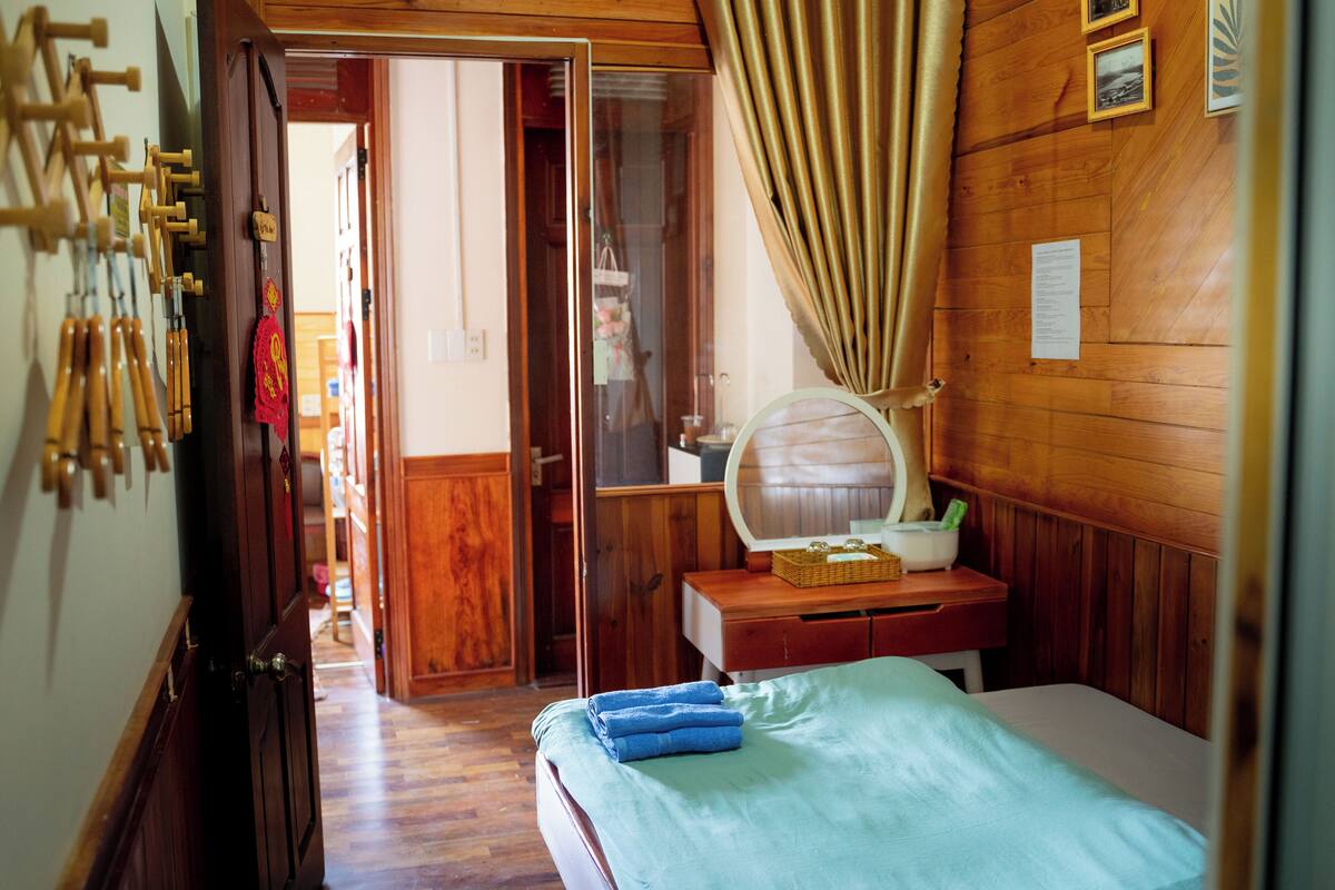 Quarto de madeira com uma cama, uma mesinha de cabeceira com espelho e uma porta que dá para outro cômodo.