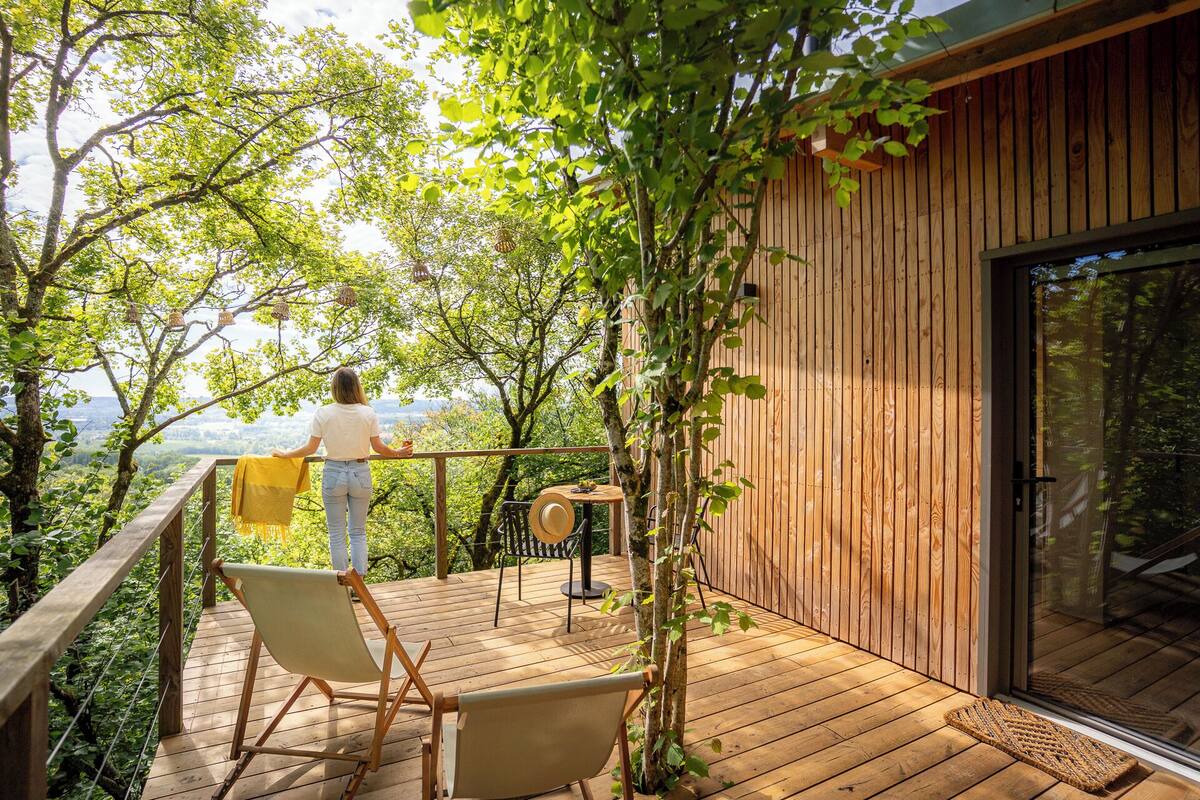 Um deck de madeira com vista para uma paisagem arborizada, uma pessoa em pé e uma mesa com uma cadeira.
