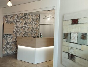 Balcão de recepção com papel de parede estampado com folhagens tropicais, luminária pendente e uma obra de arte na parede.