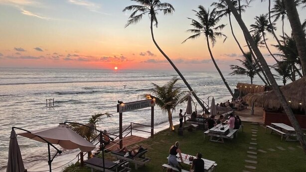 Um restaurante à beira-mar com mesas ao ar livre, palmeiras e vista para o oceano ao pôr do sol.