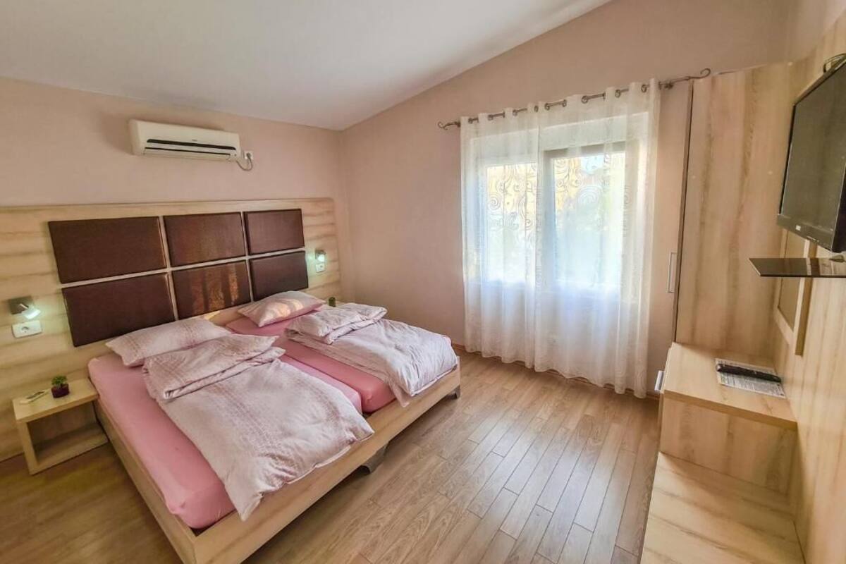 Um quarto com cama, ar condicionado, janela com cortinas e televisão em um suporte de madeira.