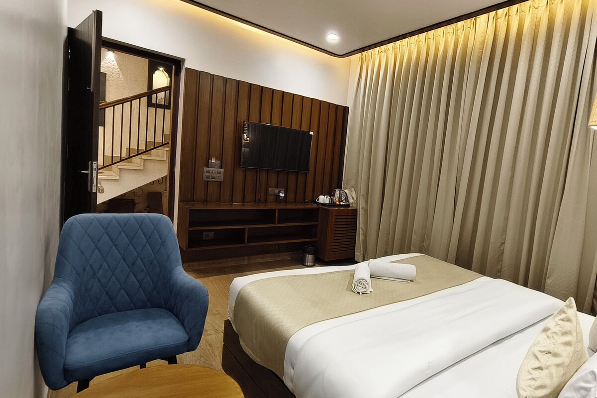 Quarto de hotel com uma cama, poltrona azul, televisão, escrivaninha com luminária e escada.