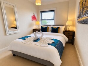 Cama bem arrumada com arranjo de toalhas em formato de coração, janela com persianas, quadro na parede e abajur na mesinha de cabeceira.