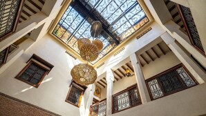Um átrio de vários níveis com grades de ferro ornamentadas, um lustre pendente no centro e vista do céu através de um claraboia.