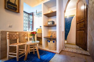 Quarto com cadeira de madeira, pequena mesa com um vaso de flores, cesta trançada e prateleira com objetos decorativos.