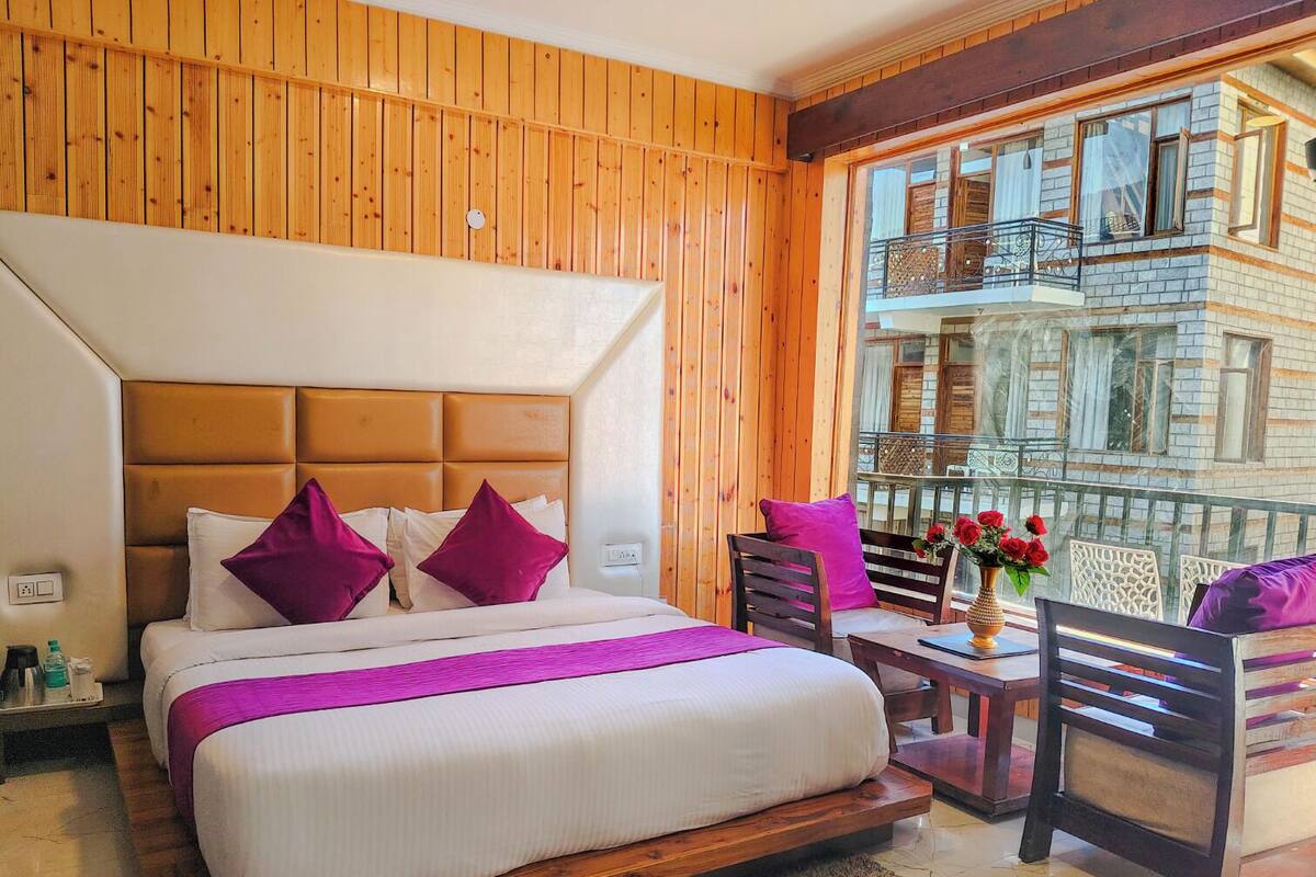 Quarto de hotel com parede revestida em madeira, uma cama grande com cabeceira, uma mesa pequena com cadeiras e vista para um edifício através de uma varanda.