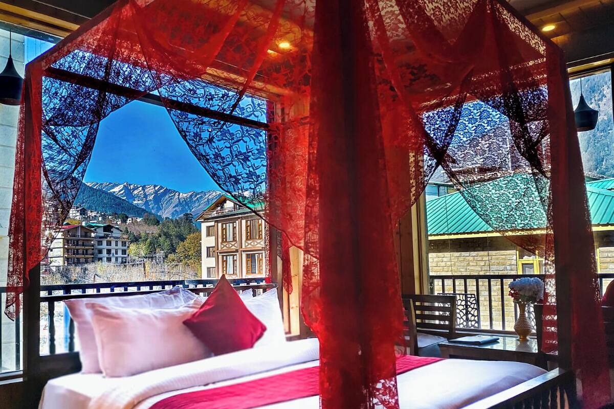 Uma cama com dossel e cortina vermelha, vista para montanhas e uma varanda com grade.
