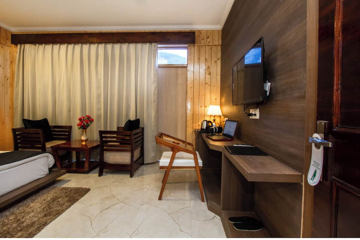Quarto de hotel com cama, escrivaninha com laptop, cadeira, mesa pequena com um vaso de flores, janela com cortinas e televisão na parede.