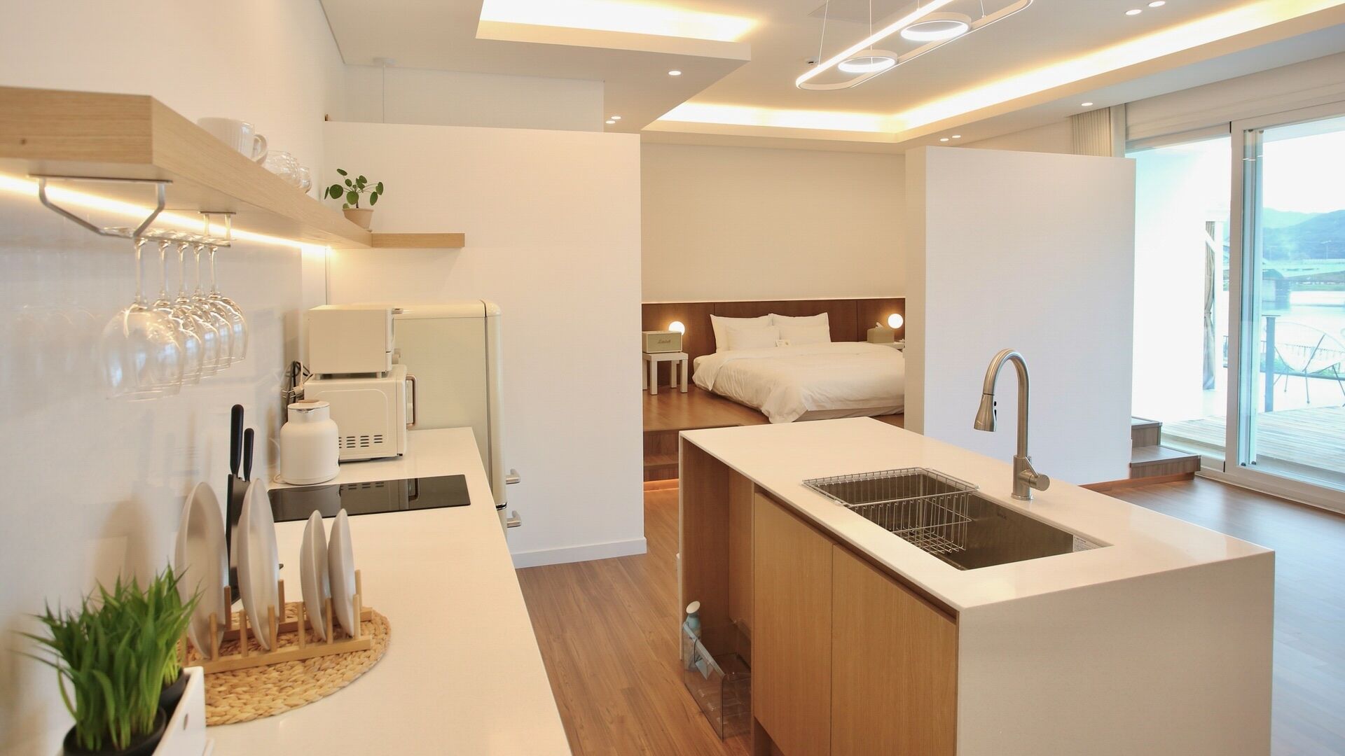 Quarto de hotel moderno com cozinha compacta, cama com encosto e vista através de uma porta de vidro.