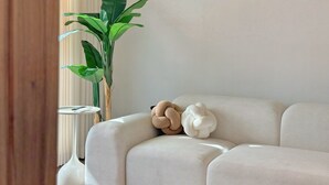 Canto de um quarto com um sofá branco, uma planta em vaso e uma pequena mesa branca com um controle remoto.