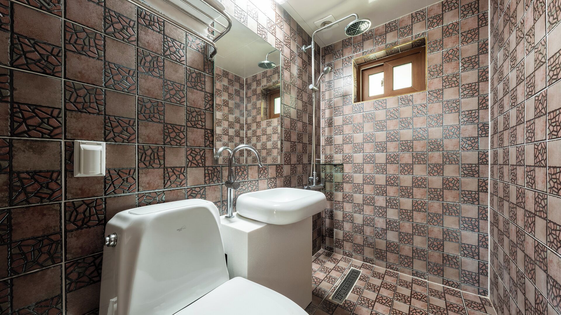 Casa de banho com sanita, lavatório, chuveiro e paredes revestidas de azulejos decorativos.