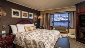 Quarto de hotel com cama king size, televisão, sofá, janela grande com vista para a água e obras de arte na parede.