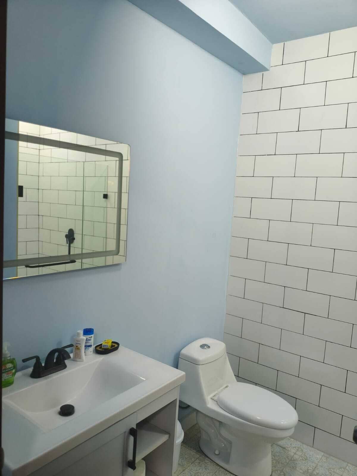 Casa de banho com lavatório branco, espelho e sanita, e parede revestida a azulejos.