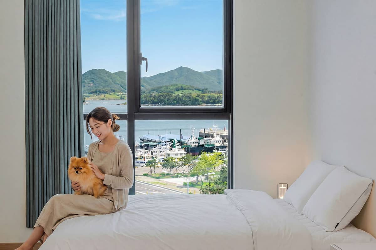 Quarto com cama, janela com vista para um corpo de água e colinas, e uma pessoa sentada na cama segurando um cão.