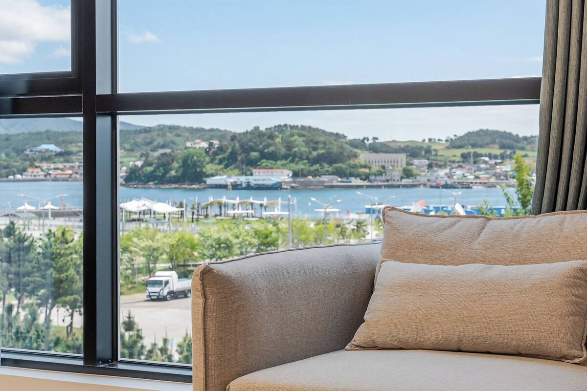 Quarto com uma grande janela que oferece vista para uma marina, barcos e uma paisagem montanhosa.