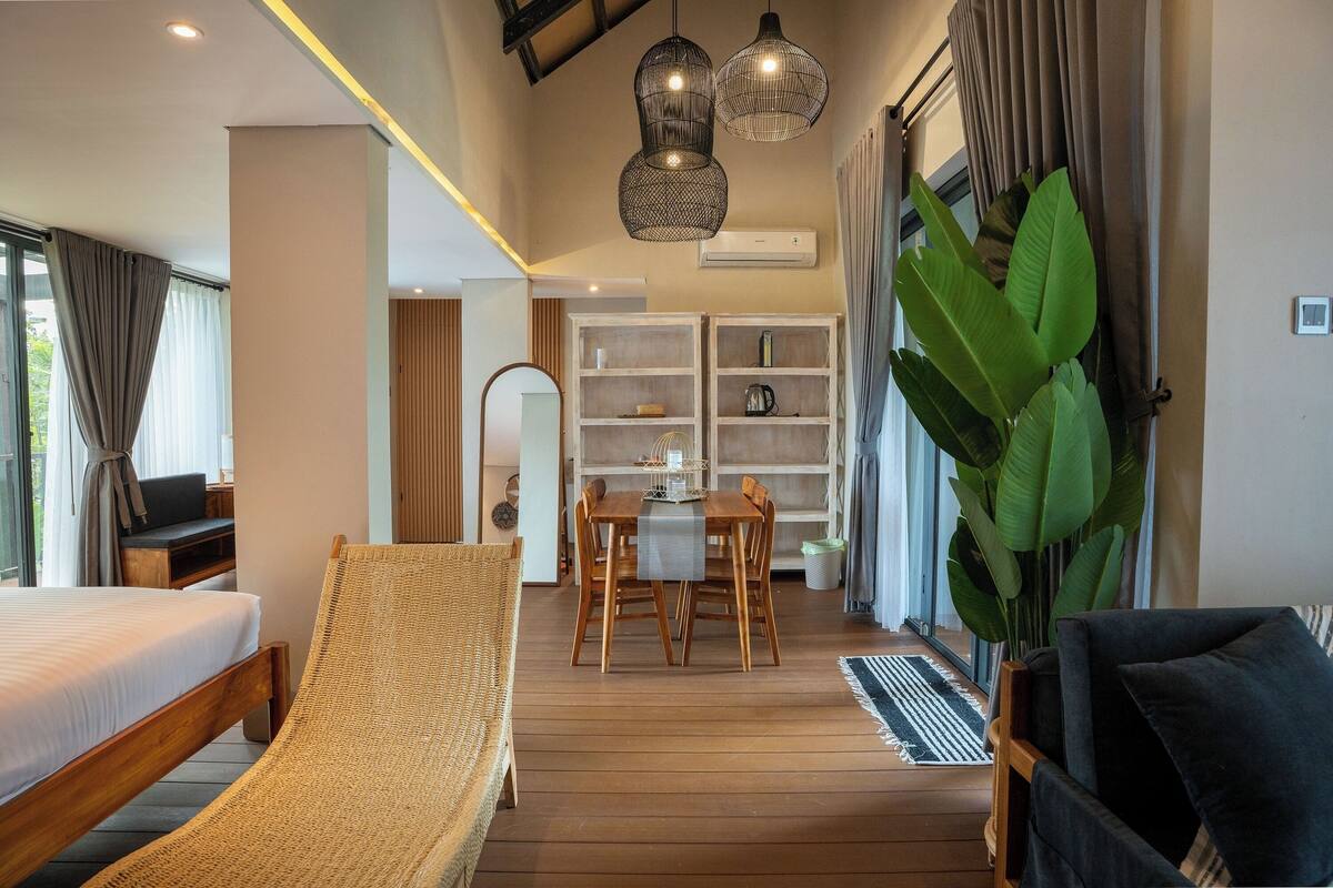 Quarto de hotel com uma cama, uma cadeira de vime, uma mesa de jantar com cadeiras, uma planta grande e uma estante de livros.