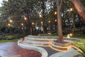 Área de bancos de concreto com curvas, iluminada por luzes de corda, cercada por árvores e vegetação.