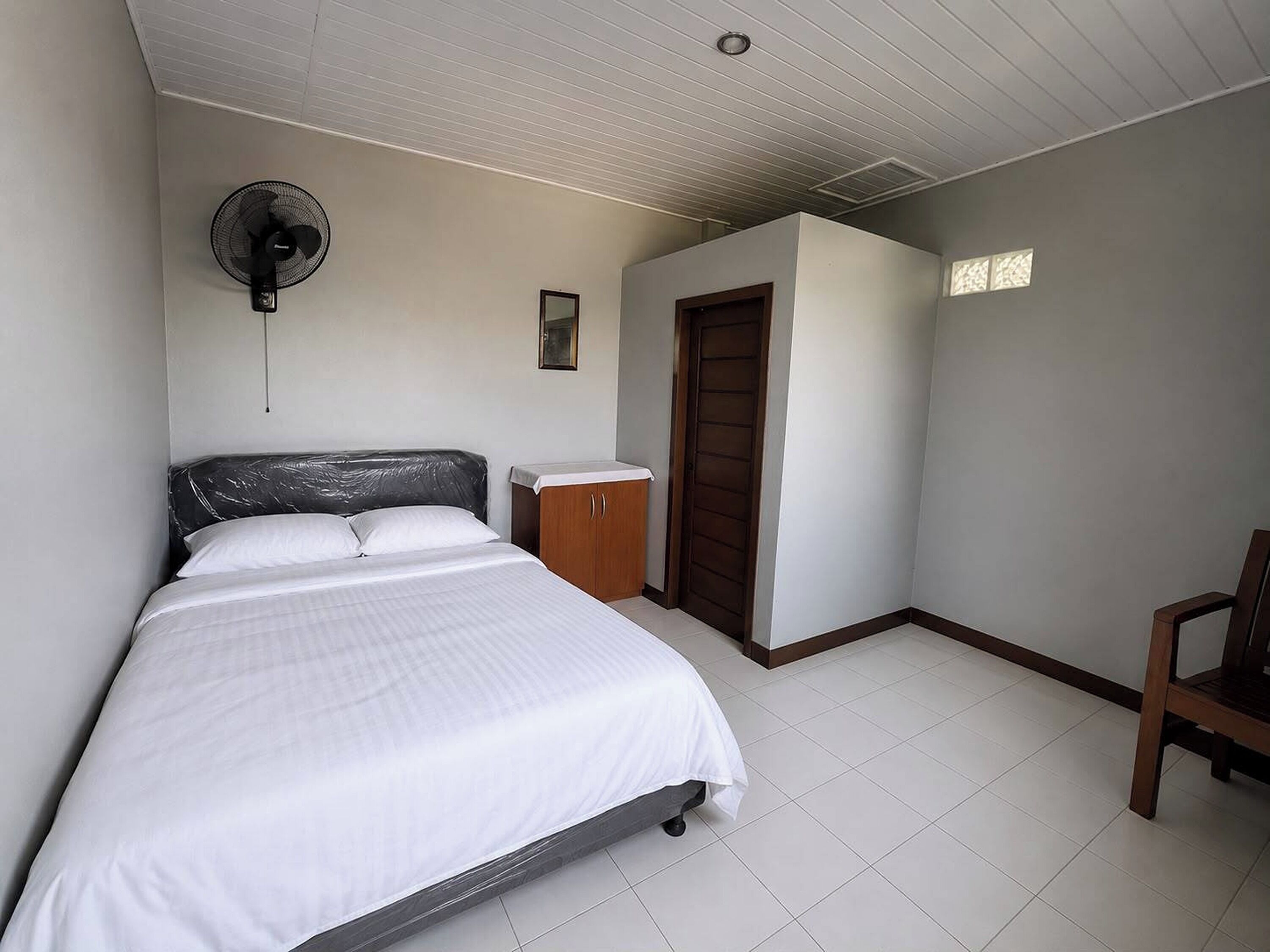 Quarto com cama, guarda-roupa de madeira, porta, ventoinha e uma pequena imagem na parede.