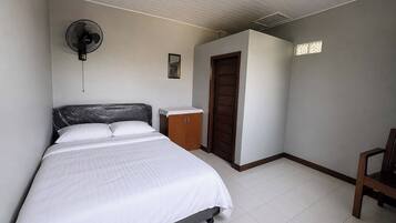 Quarto com cama, guarda-roupa de madeira, porta, ventoinha e uma pequena imagem na parede.