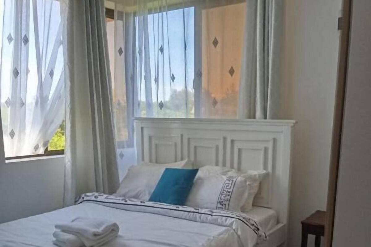 Uma cama bem arrumada com lençóis brancos e um travesseiro azul, cabeceira branca e um tapete azul sobre piso de azulejos.