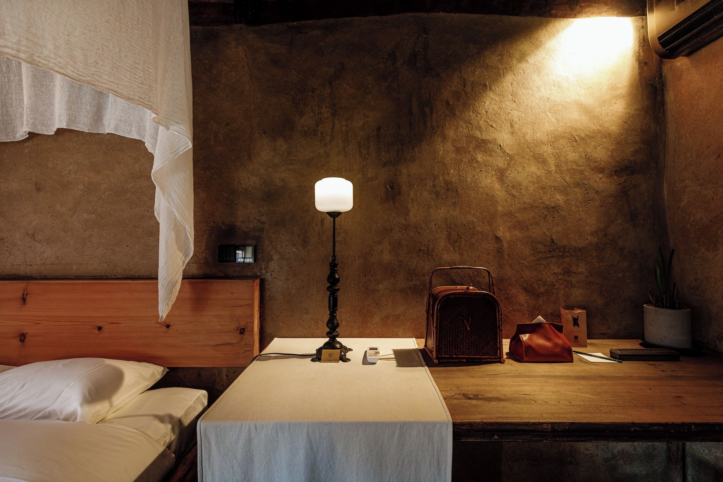Quarto com uma cama, mesa de cabeceira com luminária, mala e uma planta.