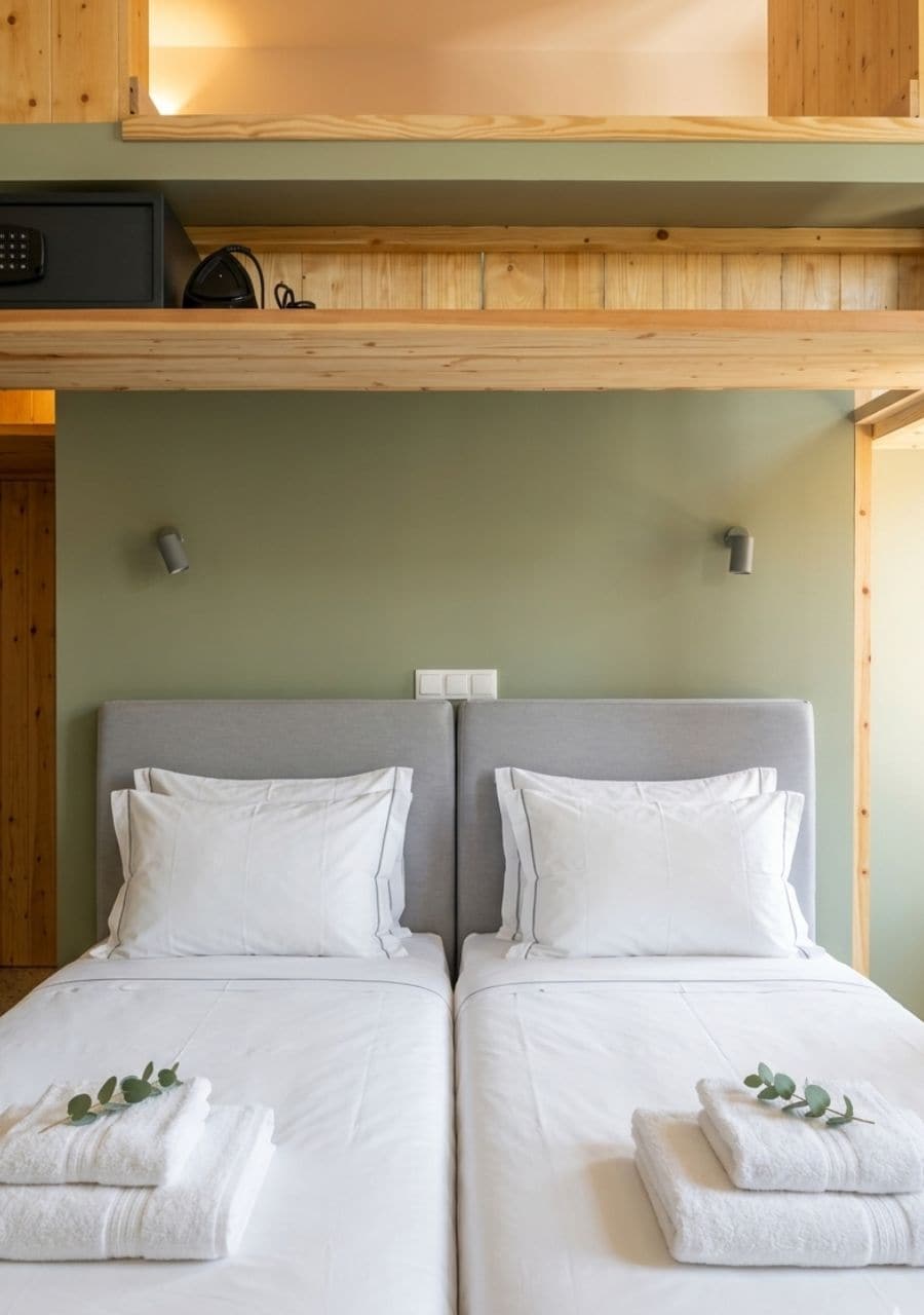 Cama de casal com roupa de cama e toalhas brancas, cabeceira de madeira e parede esverdeada.