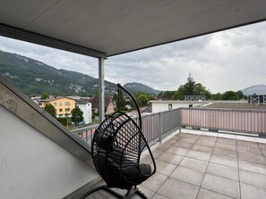 Uma varanda com uma cadeira preta, piso de azulejos e vista para uma cidade e montanhas.