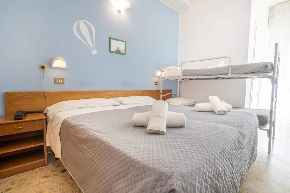Quarto de hotel com uma cama beliche, mesa de cabeceira com telefone, abajur e parede decorada com um balão de ar quente e um quadro.