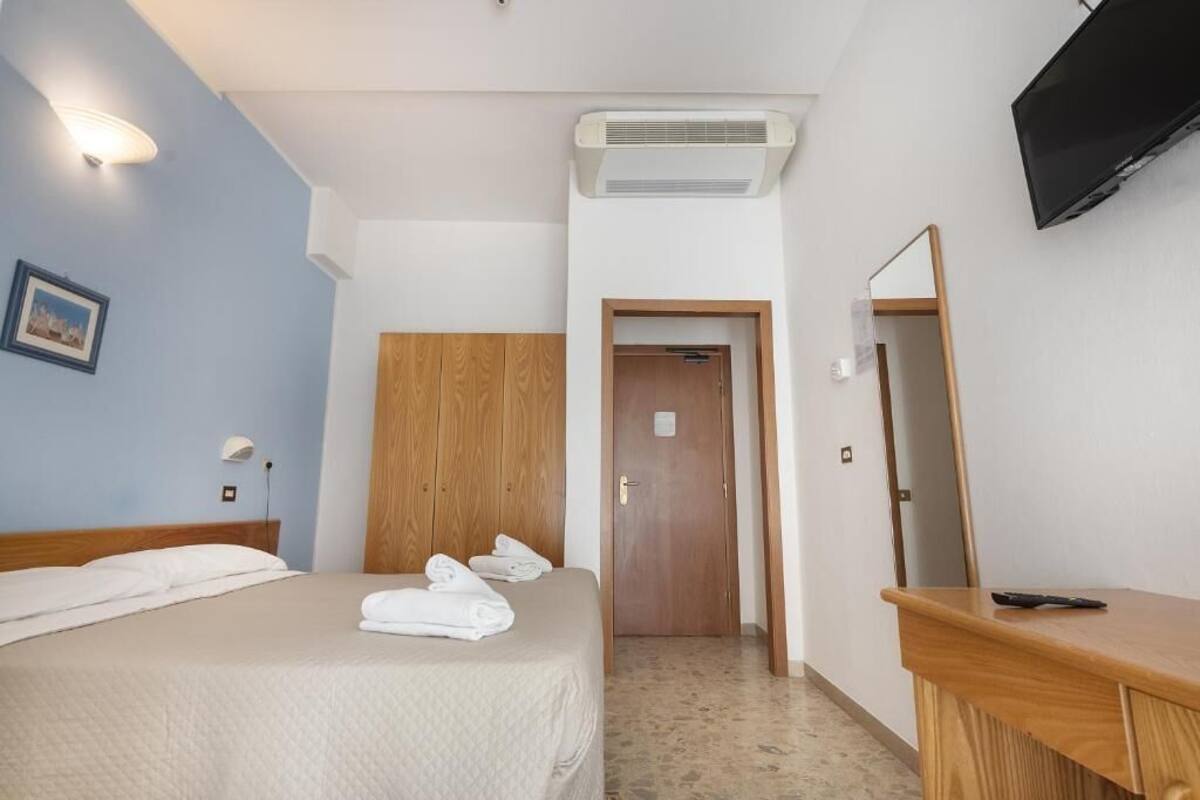 Quarto de hotel com cama, guarda-roupa de madeira, espelho, televisão e ar condicionado.