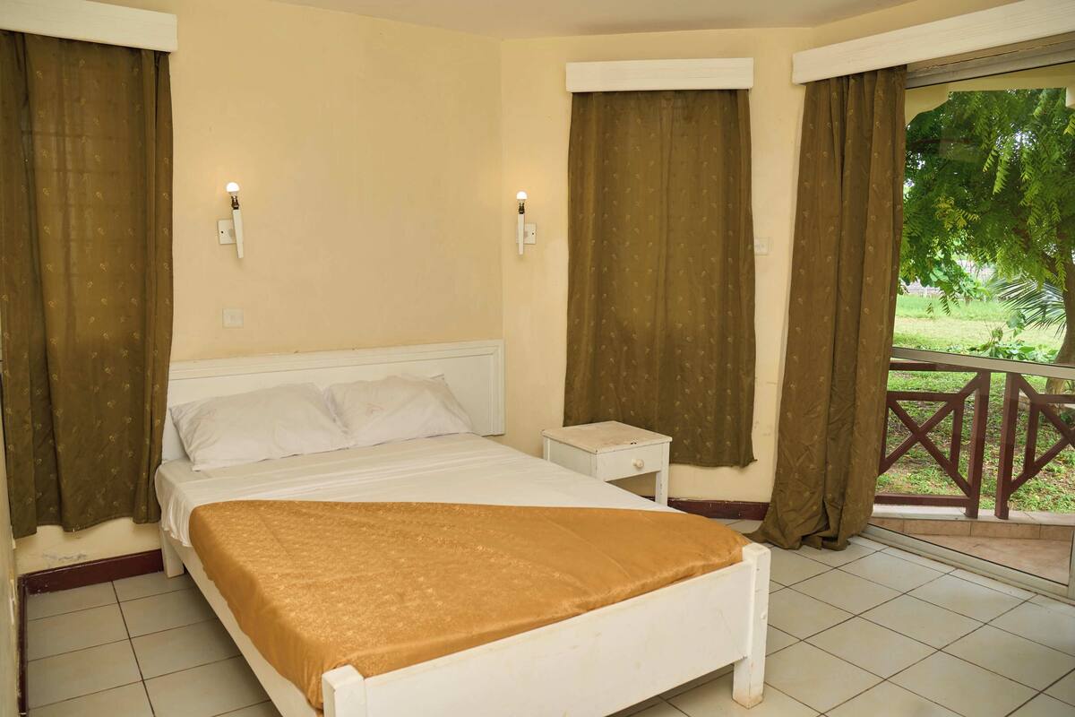 Quarto com cama, mesa de cabeceira, cortinas e vista para o verde lá fora.