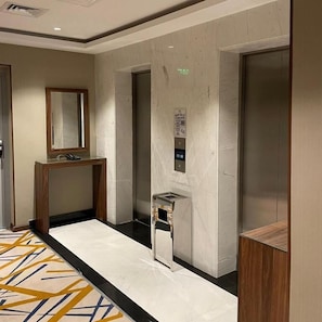 Um elevador moderno com painel digital, parede de mármore e mesa consola de madeira.
