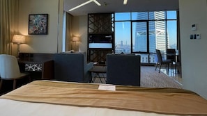 Quarto de hotel com cama, escrivaninha, duas cadeiras, televisão e uma janela grande com vista para a cidade.